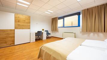 Quarto de hotel com parede de destaque em madeira, uma mesa com cadeira, cama com colcha bege e janela com cortinas.
