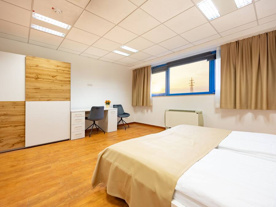 Quarto de hotel com parede de destaque em madeira, uma mesa com cadeira, cama com colcha bege e janela com cortinas.