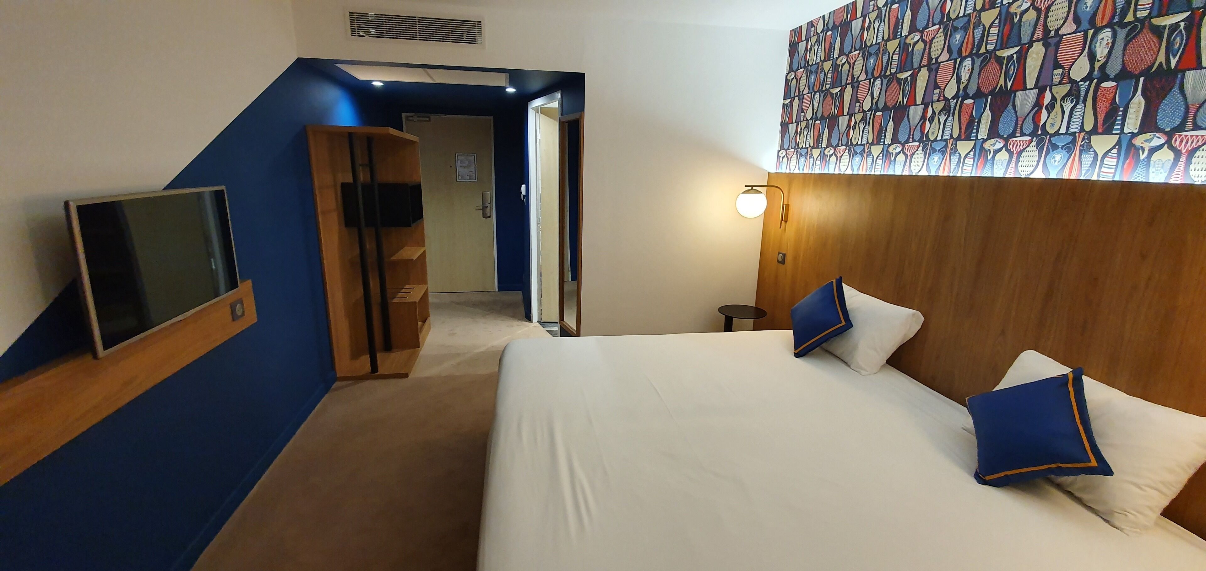 Quarto de hotel com uma cama, televisão, guarda-roupa de madeira, porta que dá para outro espaço e parede com um padrão decorativo.