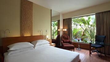 Quarto de hotel com uma cama grande, uma escrivaninha com cadeira, uma mesinha e uma janela com vista para plantas tropicais.