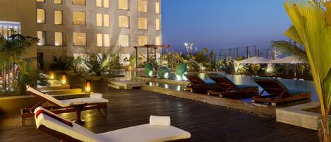 Área de piscina no terraço com espreguiçadeiras, um elemento aquático e um edifício com janelas iluminadas.