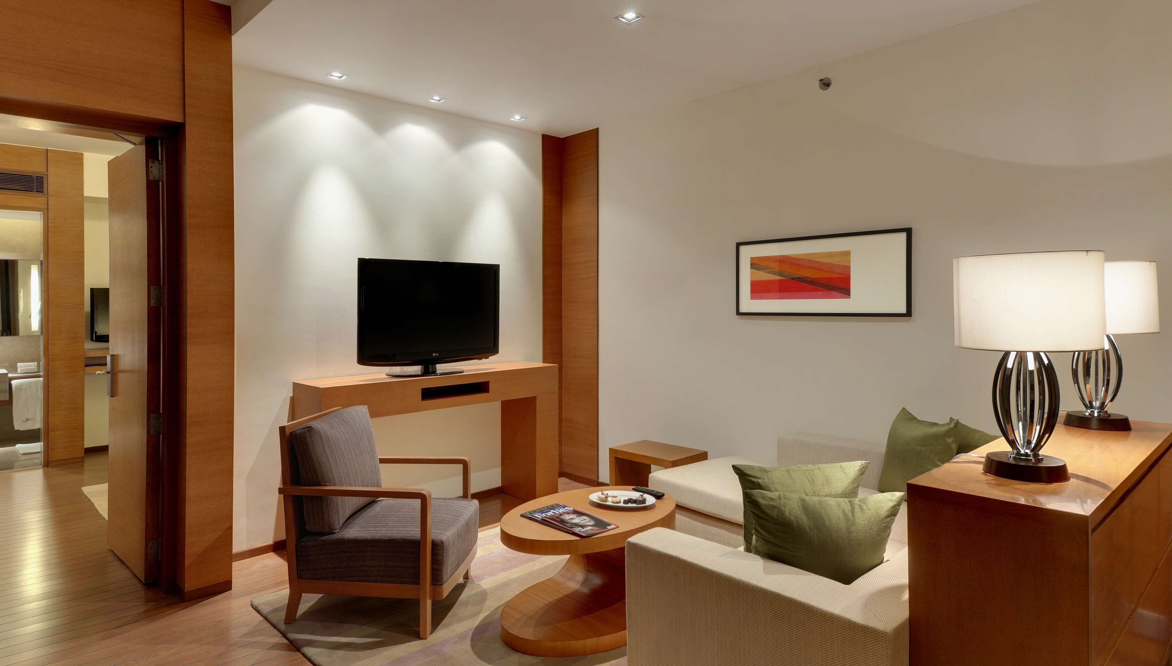 Quarto de hotel moderno com televisão de tela plana presa a um suporte de madeira, um sofá, uma poltrona, uma mesa de centro e uma mesinha de cabeceira com abajur.