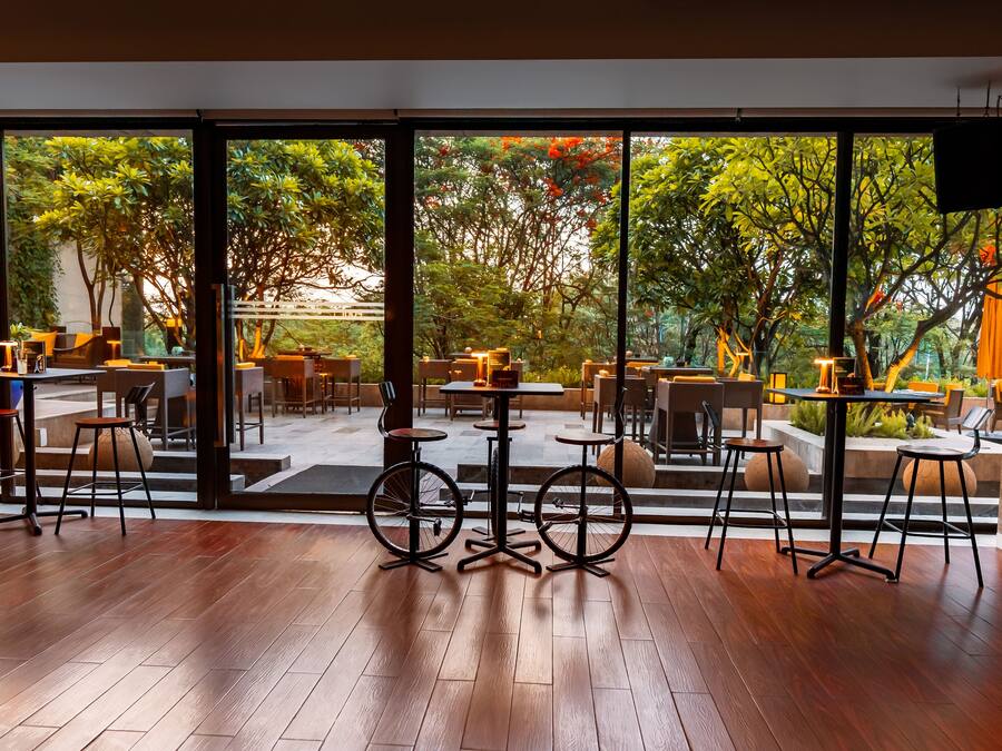 Espaço interno com assentos, chão de madeira, mesas e cadeiras, grandes janelas com vista para vegetação e árvores.