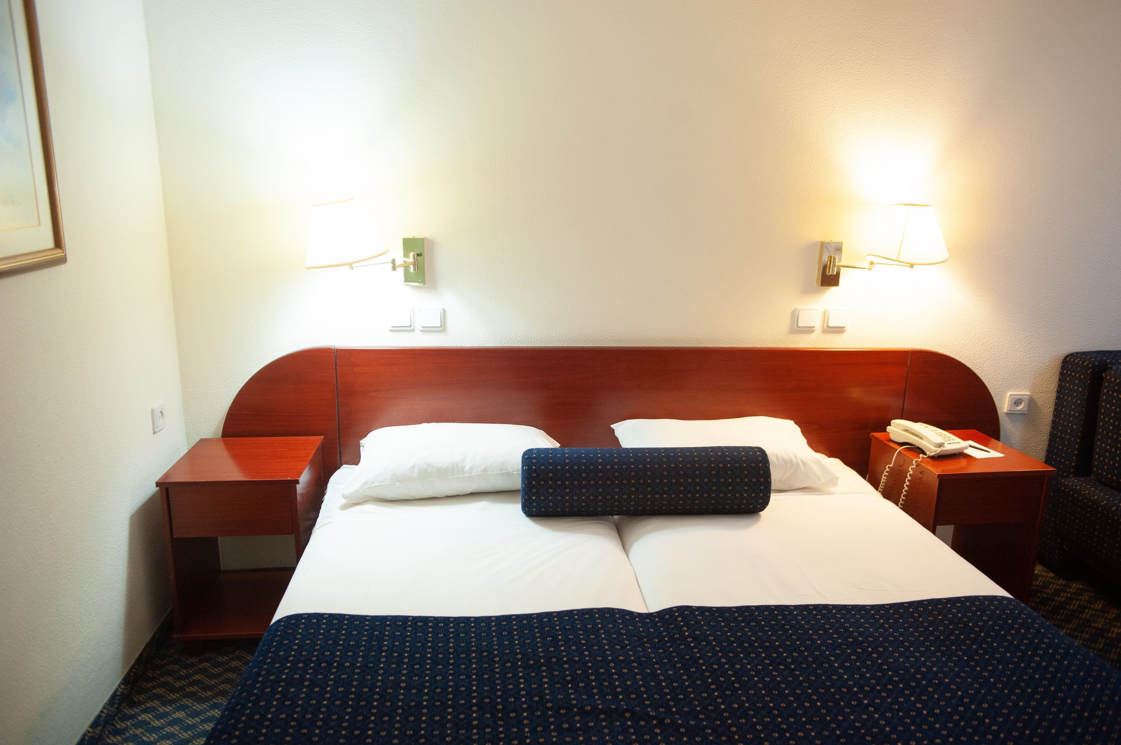 Quarto de hotel com uma cama de casal, duas mesas de cabeceira, uma luminária e uma cadeira, além de duas luminárias fixadas na parede.