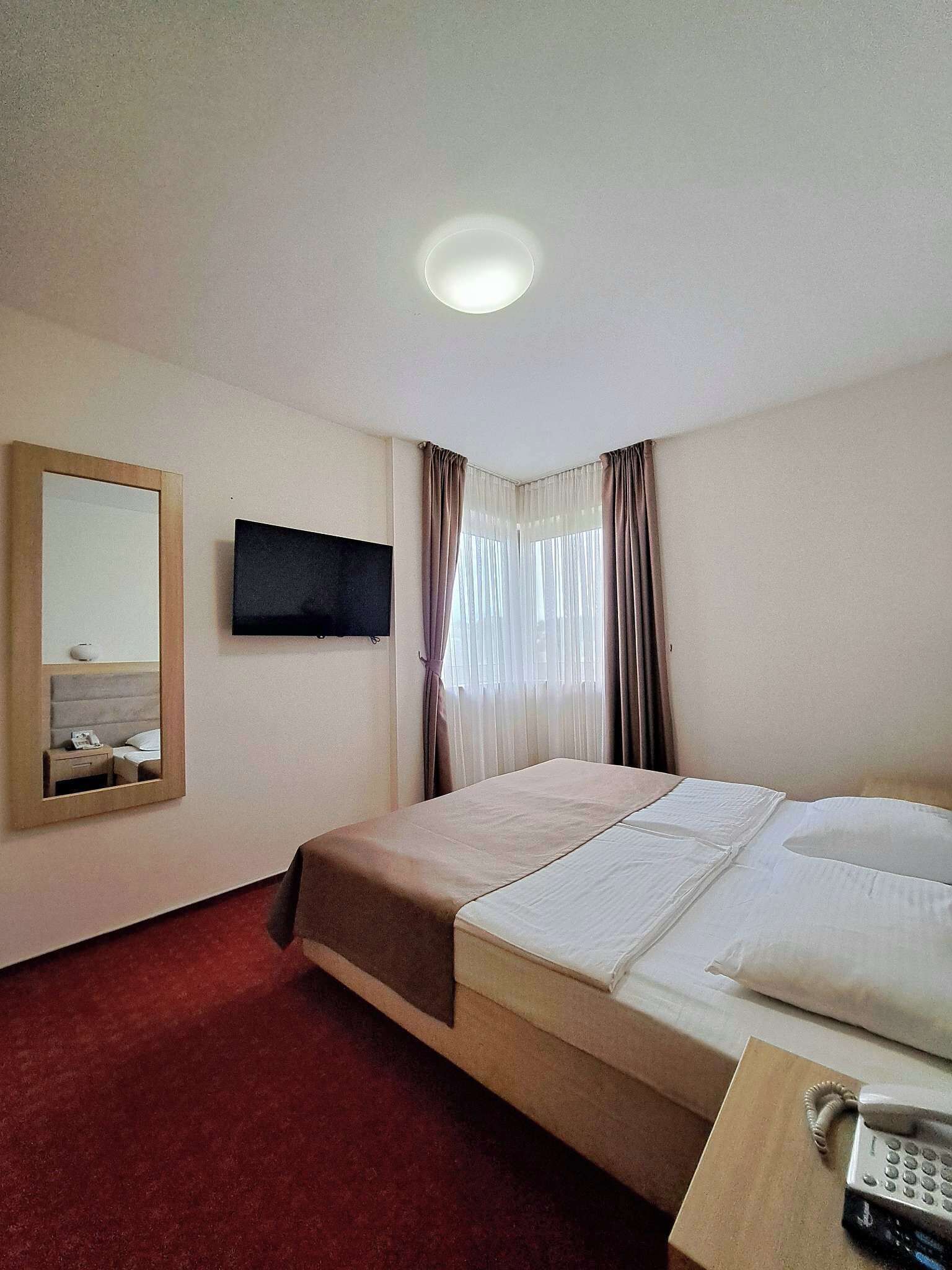 Quarto de hotel com cama, telefone ao lado da cama, televisão presa na parede e um espelho refletindo parte do quarto.