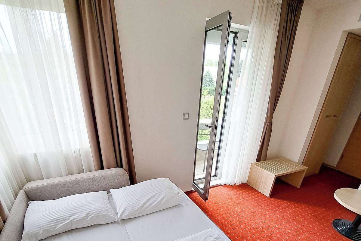 Quarto de hotel com uma cama, uma pequena mesa, uma cadeira e vista para o exterior através de uma porta de vidro.