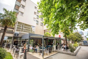 Uma área de café ao ar livre com mesas e cadeiras, cercada por edifícios e vegetação.