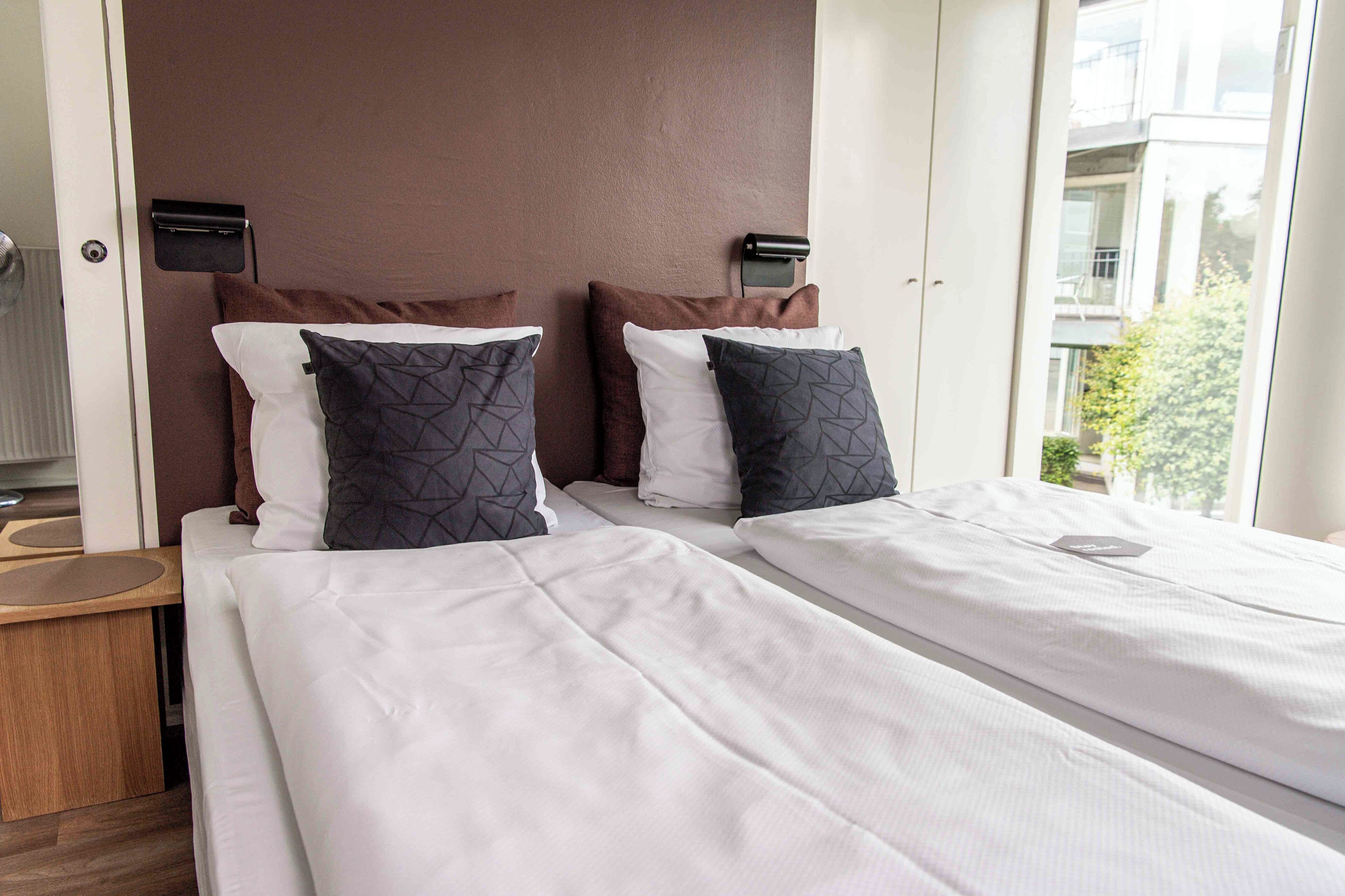 Quarto de hotel com duas camas, cada uma com roupa de cama branca e almofadas com padrão geométrico.