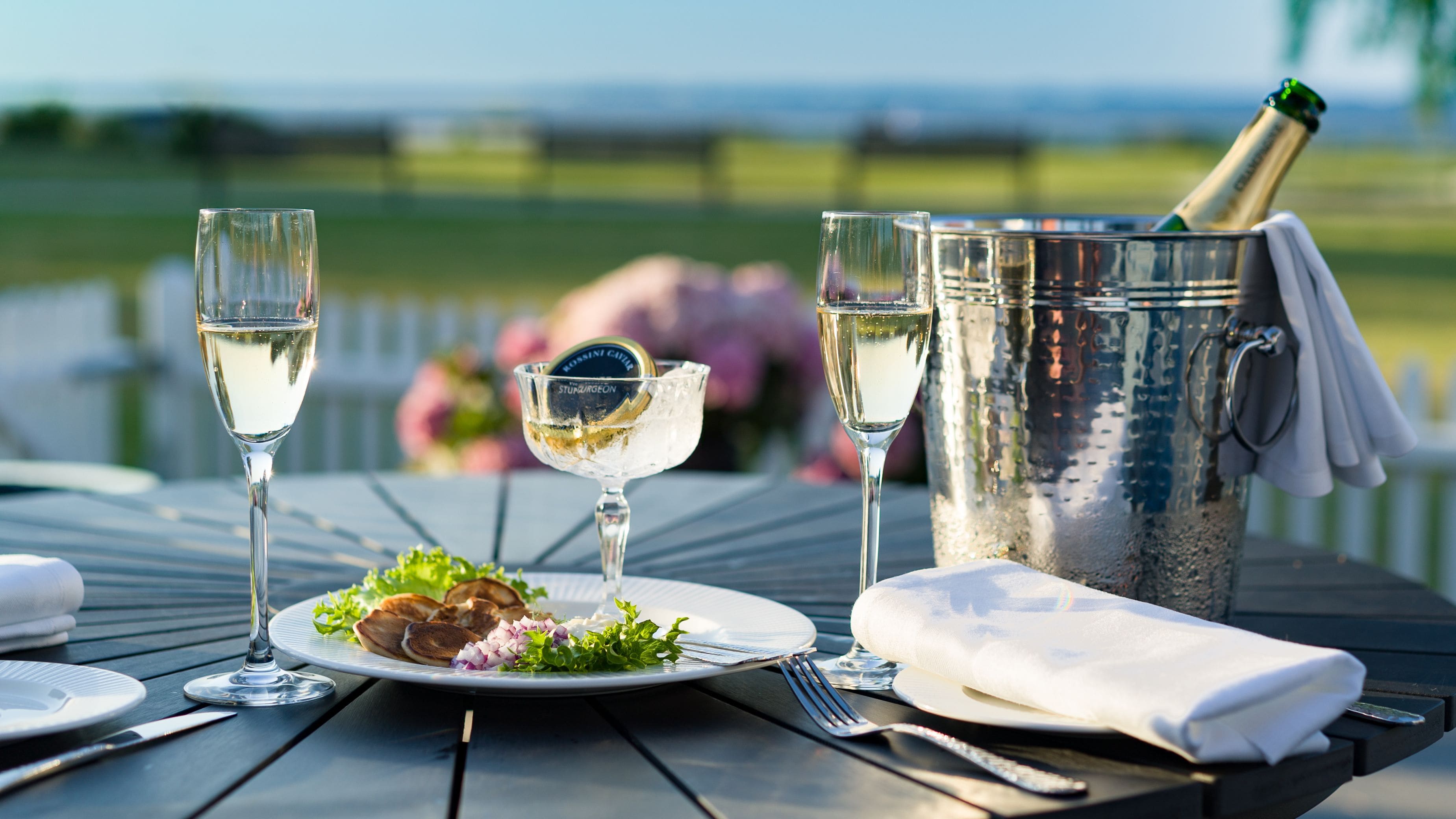 Uma mesa posta com duas taças de vinho branco, uma garrafa de champanhe, uma taça de champanhe, um prato com comida e um guardanapo.