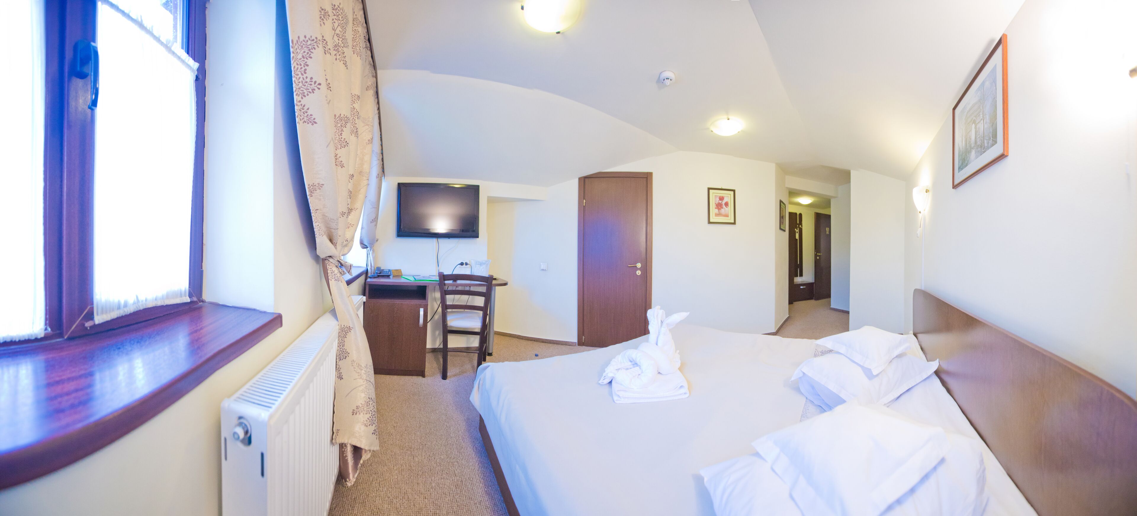 Quarto de hotel com cama, escrivaninha, cadeira, televisão e banheiro ao fundo.