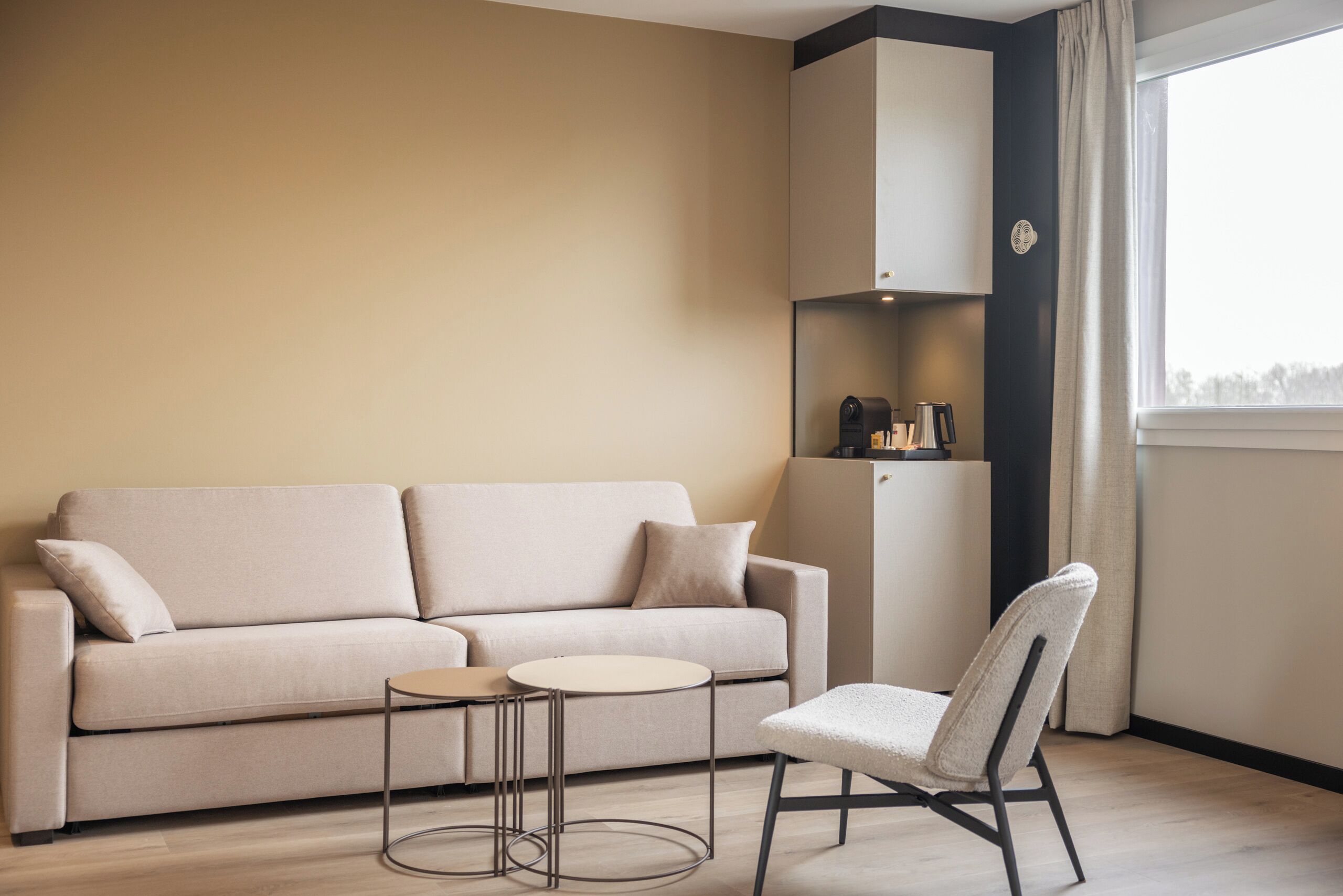 Uma sala de estar moderna com um sofá bege, uma mesa de centro redonda e uma poltrona. Há um armário com uma máquina de café e uma pequena mesa em cima.