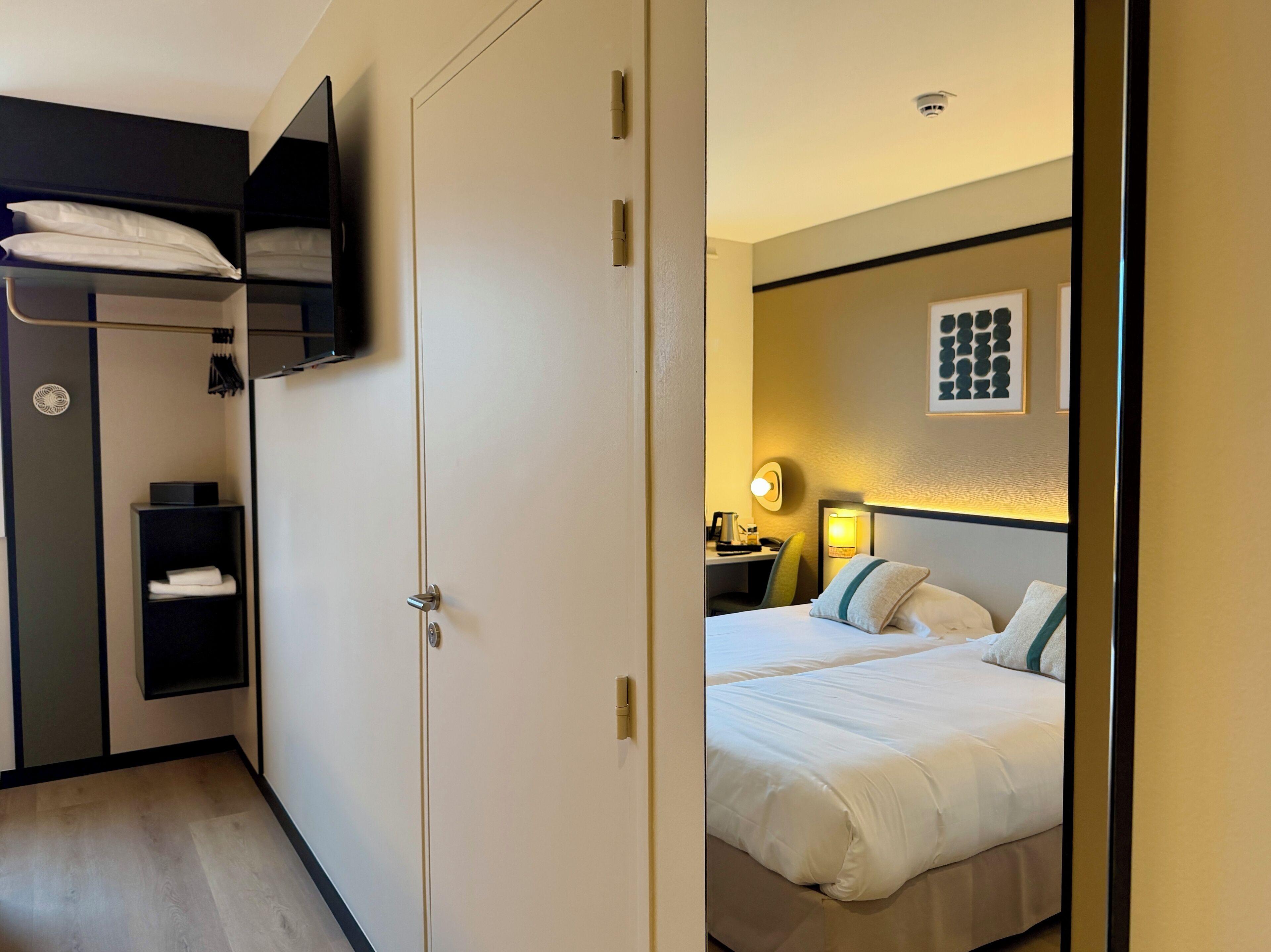 Quarto de hotel com cama, escrivaninha com luminária, prateleira com toalhas e armário.