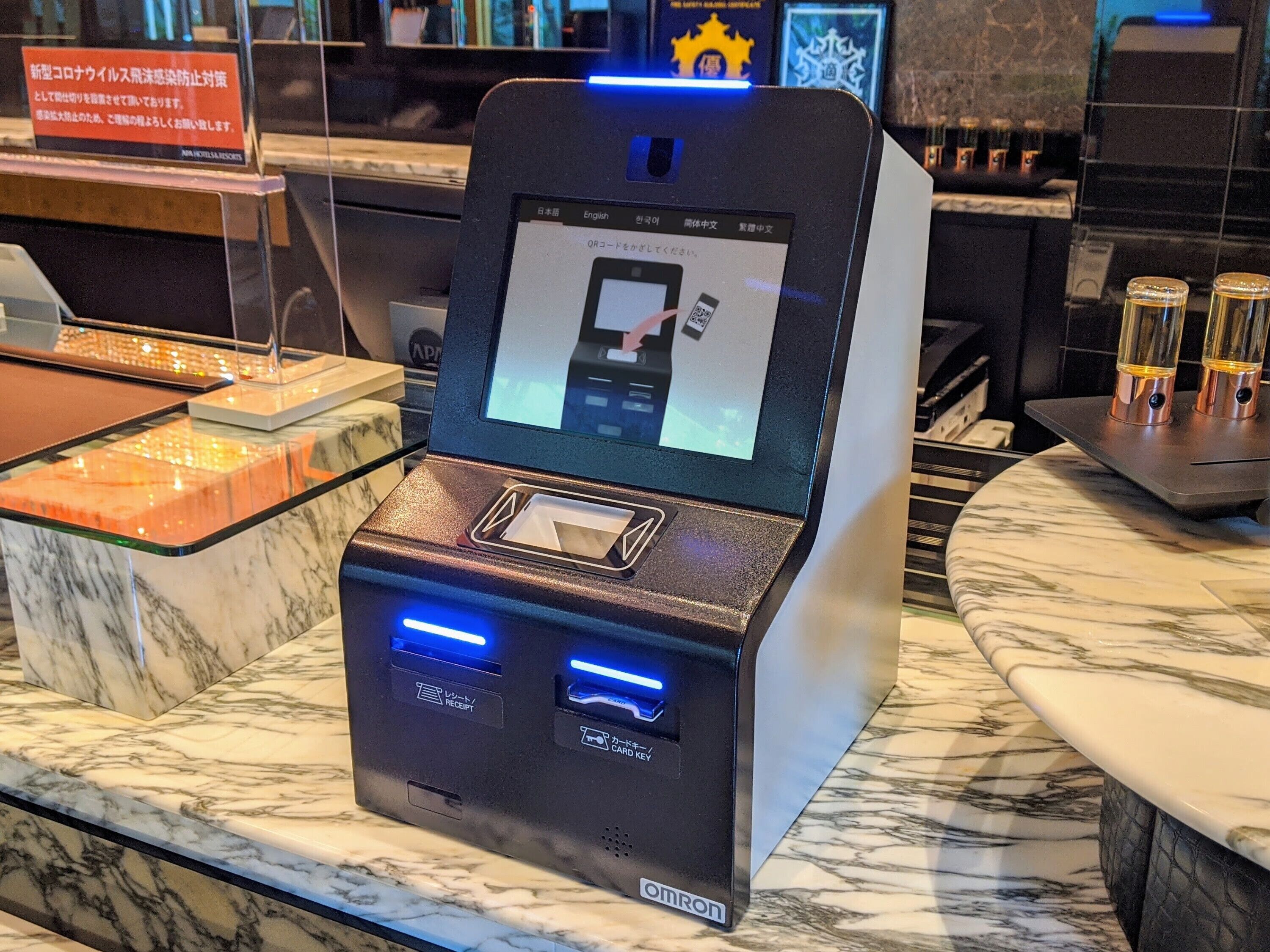 Um quiosque de autoatendimento com tela exibindo interface de celular, compartimento para recibos e leitor de cartão.