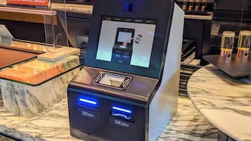 Um quiosque de autoatendimento com tela exibindo interface de celular, compartimento para recibos e leitor de cartão.