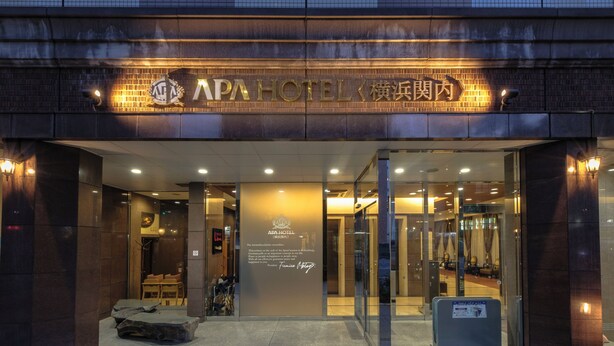 Exterior do APA HOTEL com entrada de vidro, um banco de pedra e uma placa com o nome do hotel.