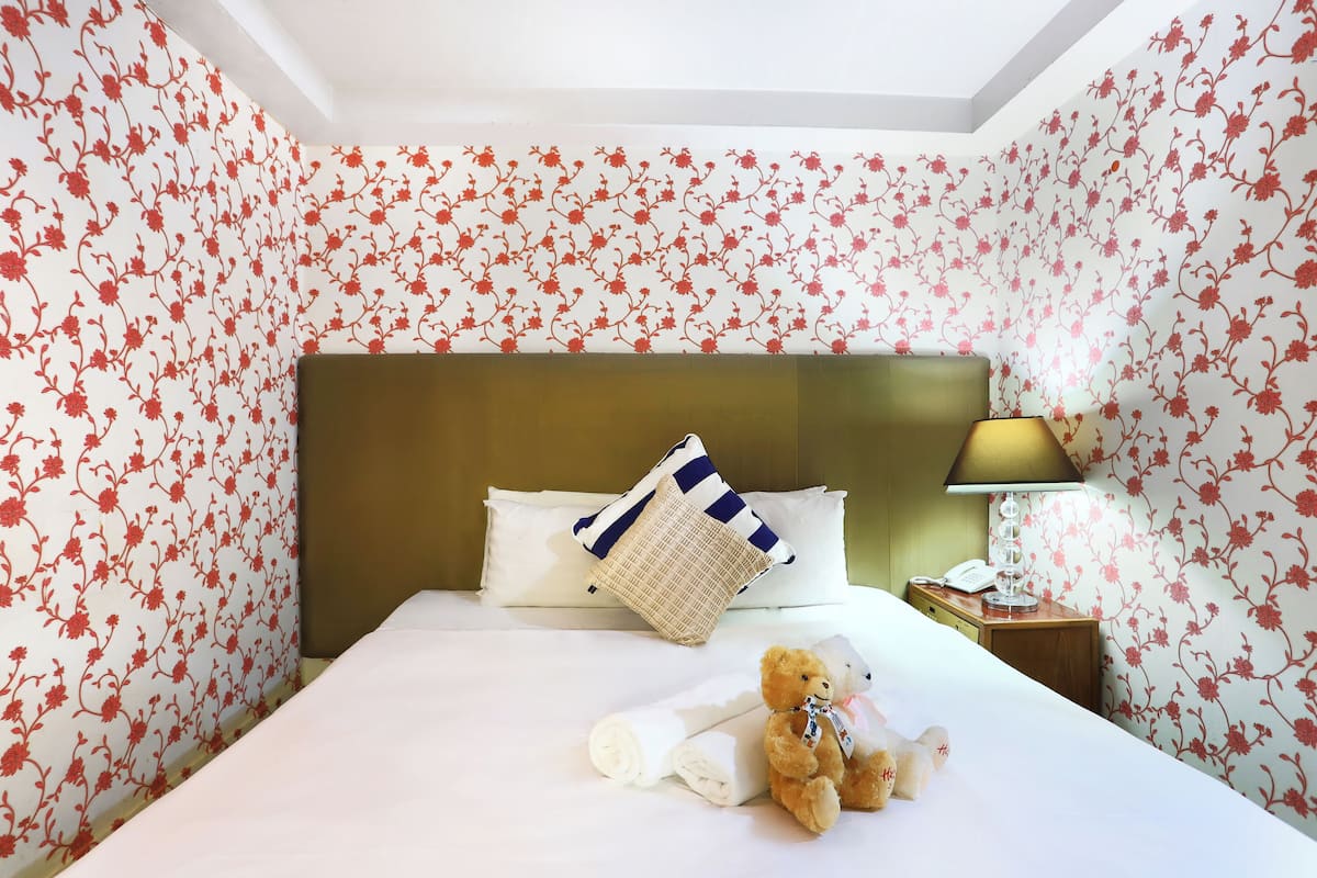Quarto de hotel com papel de parede floral, cama bem arrumada com almofadas, abajur e um ursinho de pelúcia na cama.