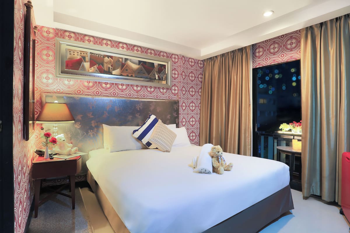 Quarto de hotel com uma cama grande, uma mesa de cabeceira com abajur, um urso de pelúcia e um espelho na parede.