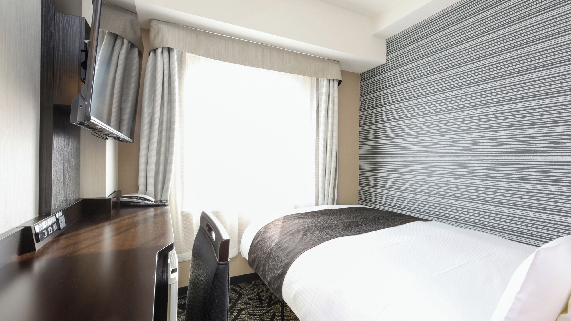 Quarto de hotel com cama, escrivaninha, cadeira e uma janela grande com cortinas.