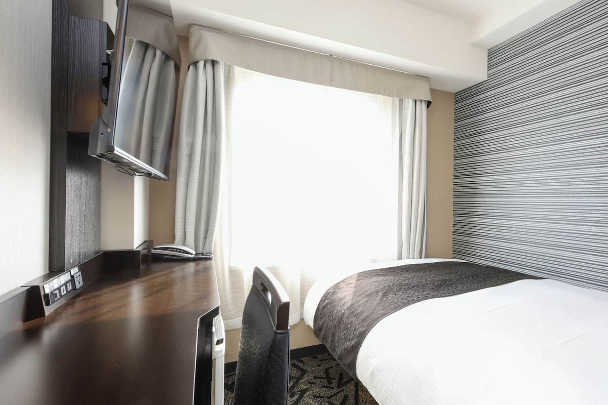 Quarto de hotel com cama, escrivaninha, cadeira e uma janela grande com cortinas.