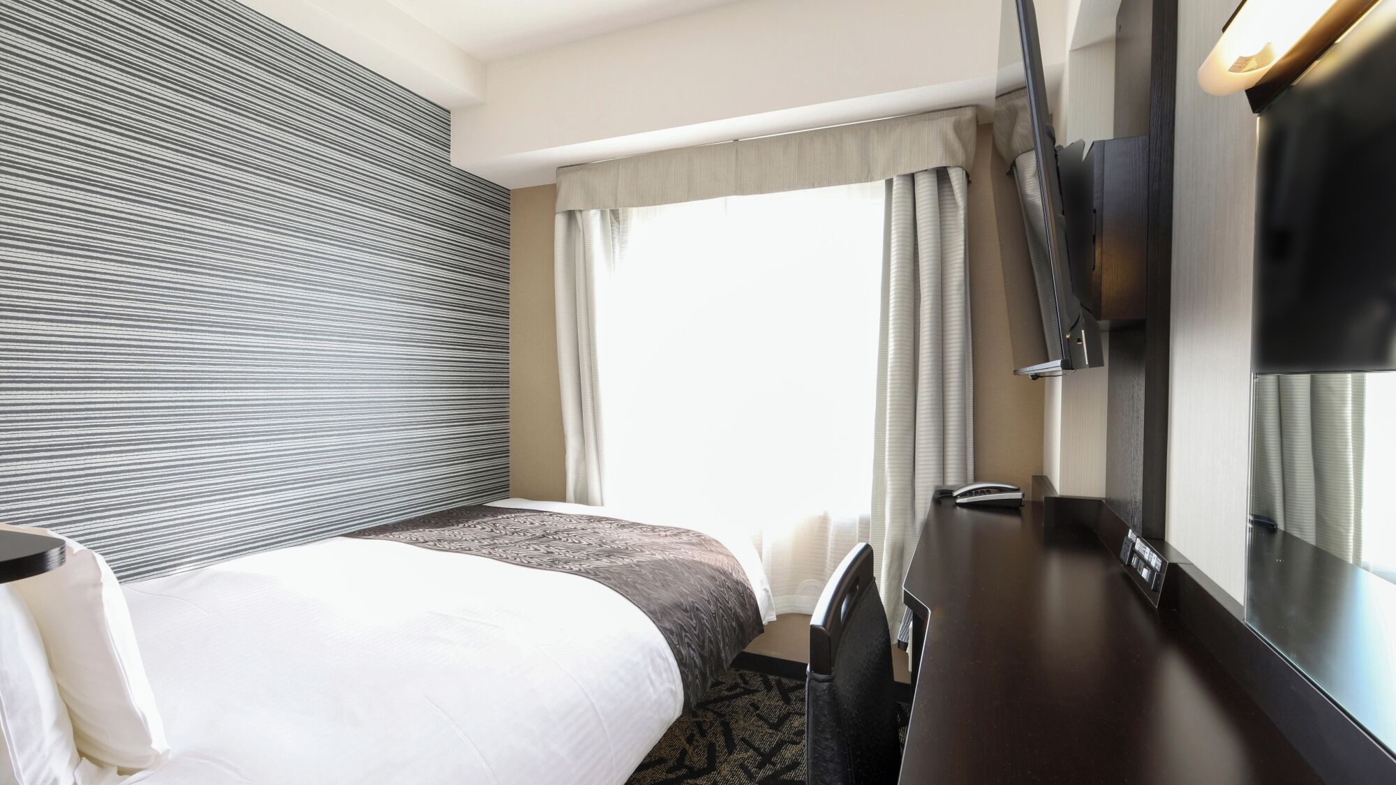 Quarto de hotel com cama, escrivaninha, televisão e janela com cortinas.
