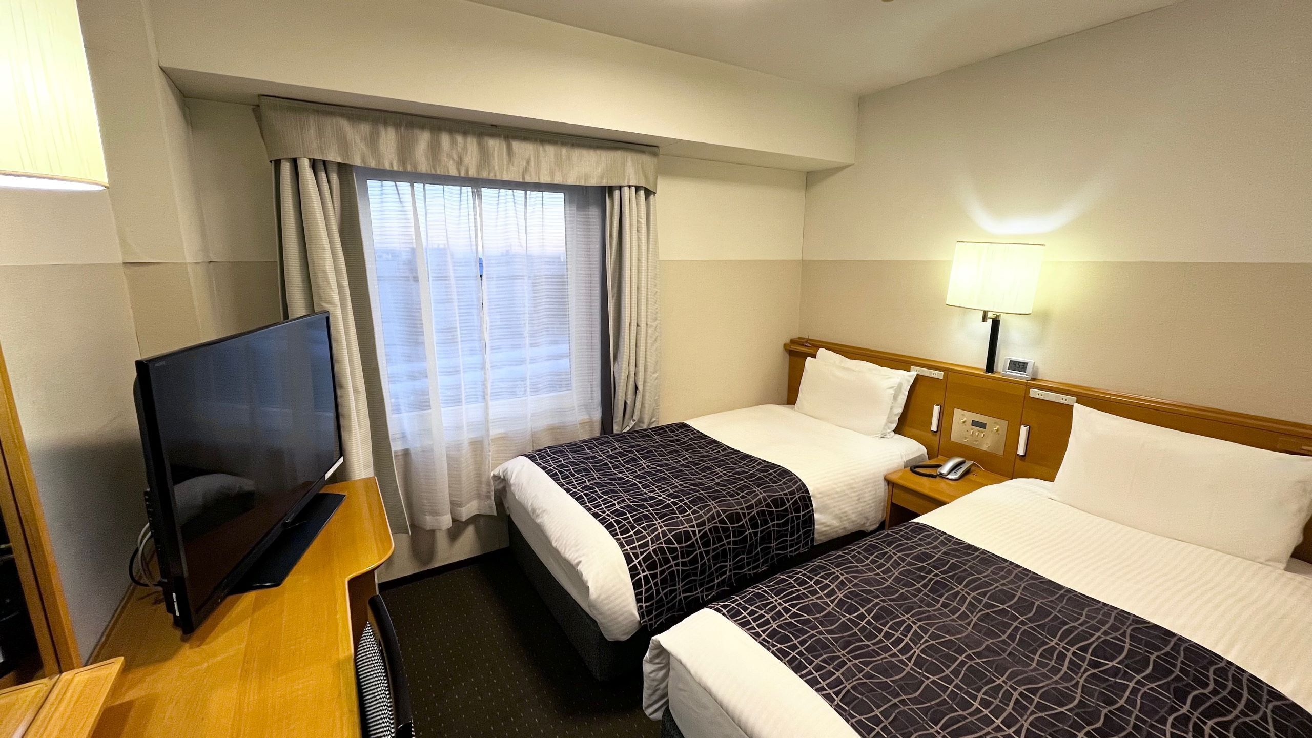 Quarto de hotel com duas camas, uma televisão, uma mesinha de cabeceira, um abajur e uma janela com cortinas.