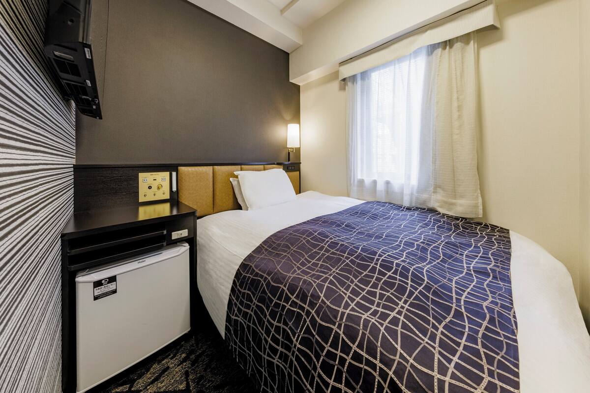 Quarto de hotel com cama, escrivaninha, minibar e janela com cortinas.