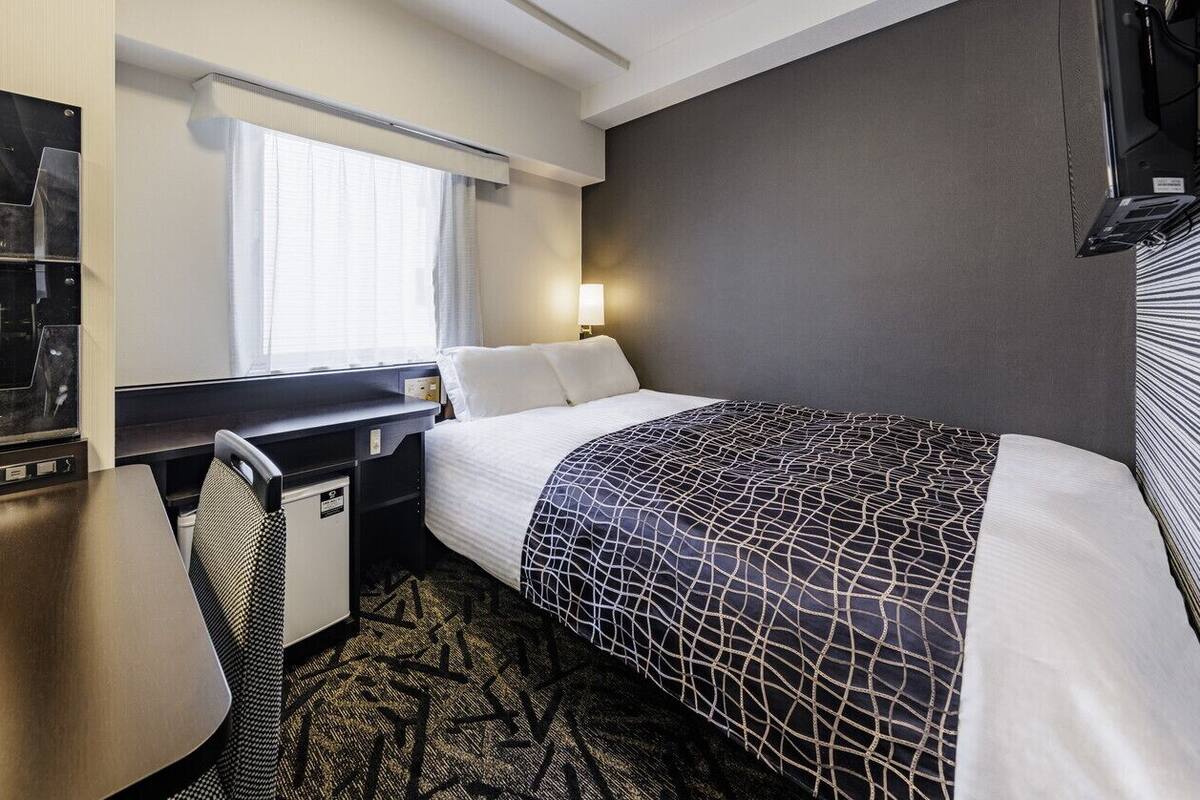 Quarto de hotel com cama, escrivaninha, cadeira e janela com cortinas.