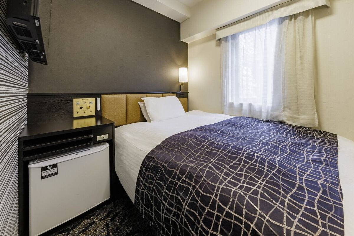 Quarto de hotel com cama, mesa de cabeceira, mini-geladeira, televisão fixada na parede e janela com cortinas.