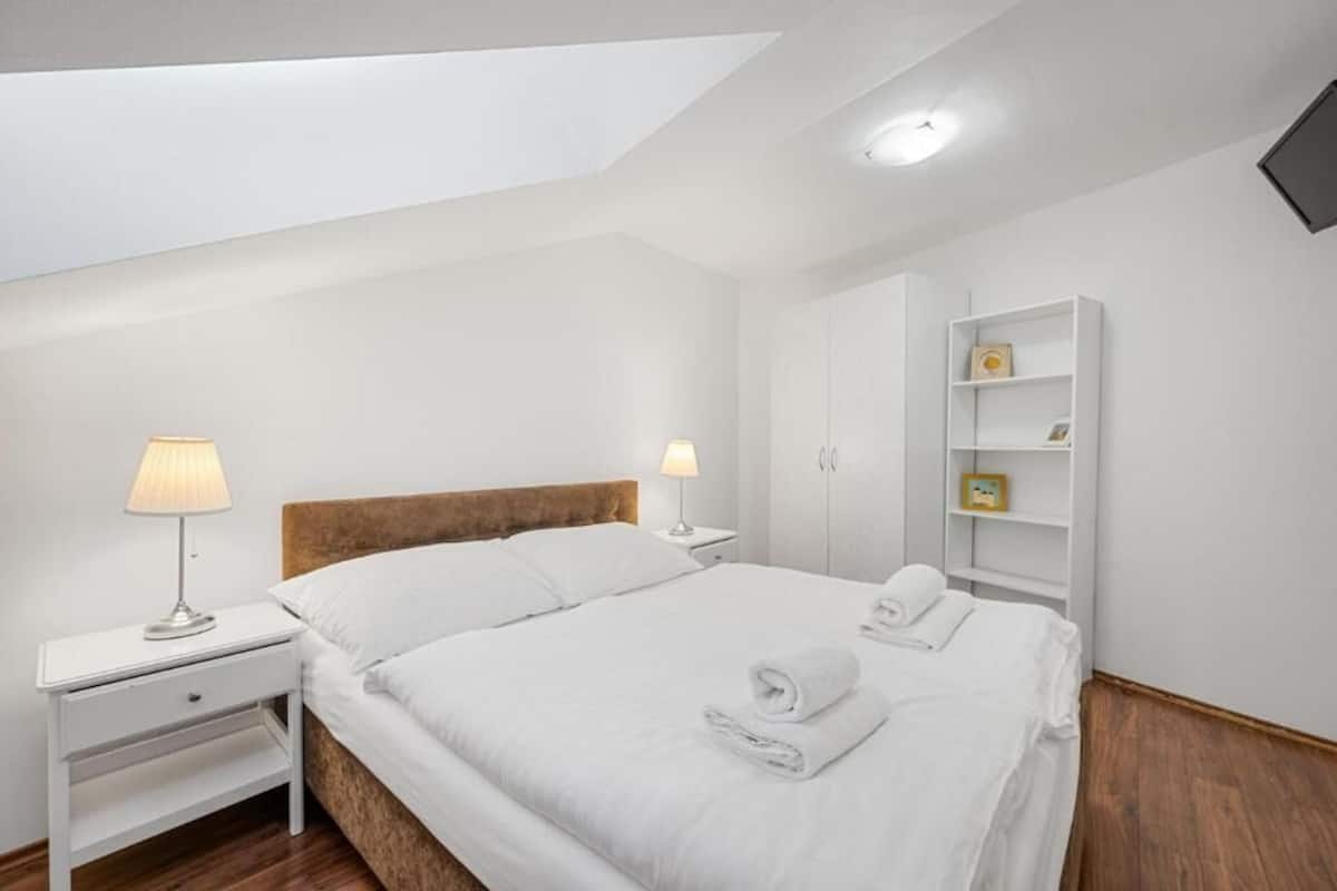 Quarto com cama, duas mesas de cabeceira, um guarda-roupa e televisão fixada na parede.
