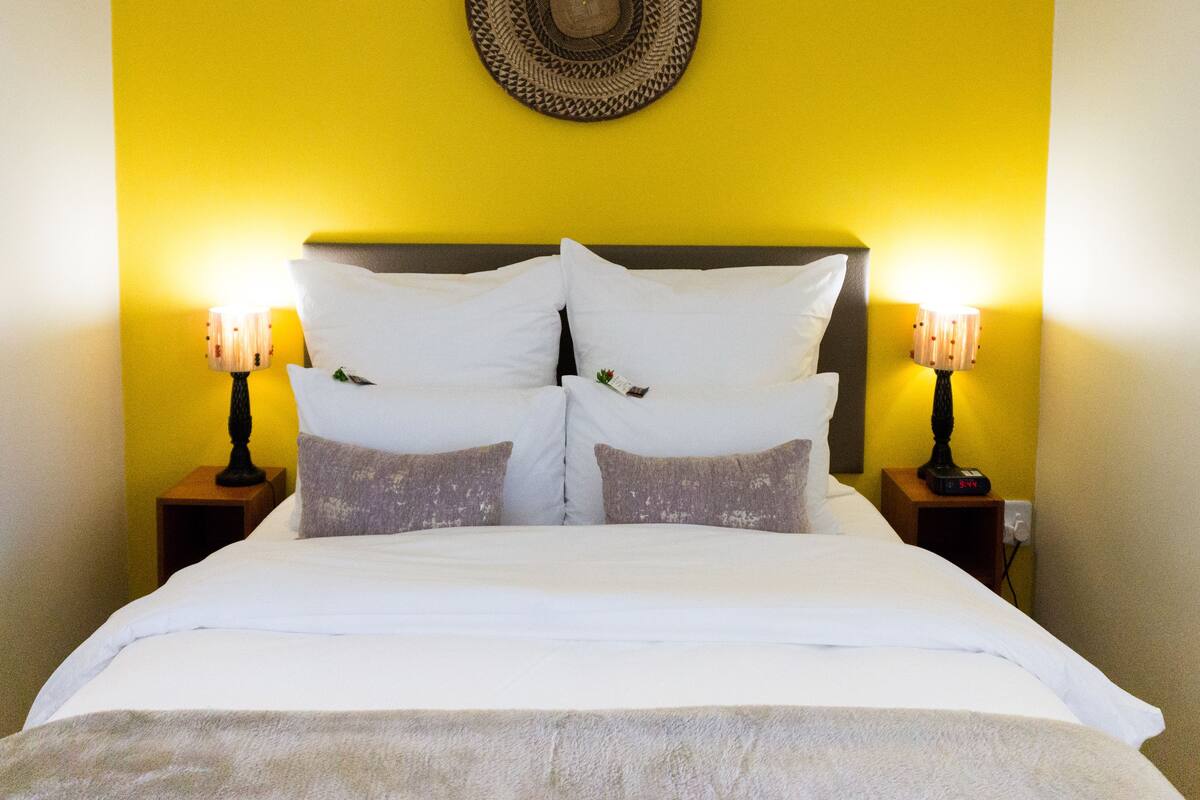 Cama com roupa de cama branca, duas mesas de cabeceira com abajures e um objeto decorativo na parede.