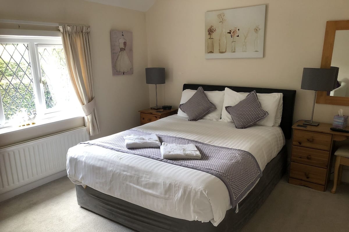 Uma cama bem arrumada com um edredom cinza e branco, duas mesas de cabeceira com abajures, uma janela com cortinas e uma parede com uma obra de arte emoldurada.