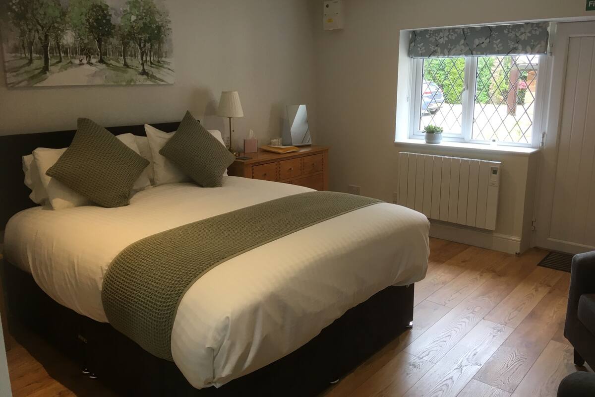 Uma cama bem arrumada com um manta verde, uma mesa de cabeceira com abajur, um radiador e uma janela com vista para o verde.