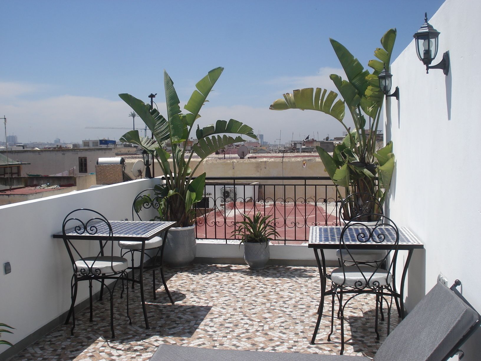 Uma varanda com duas mesas e cadeiras, plantas em vasos e vista para a paisagem urbana.
