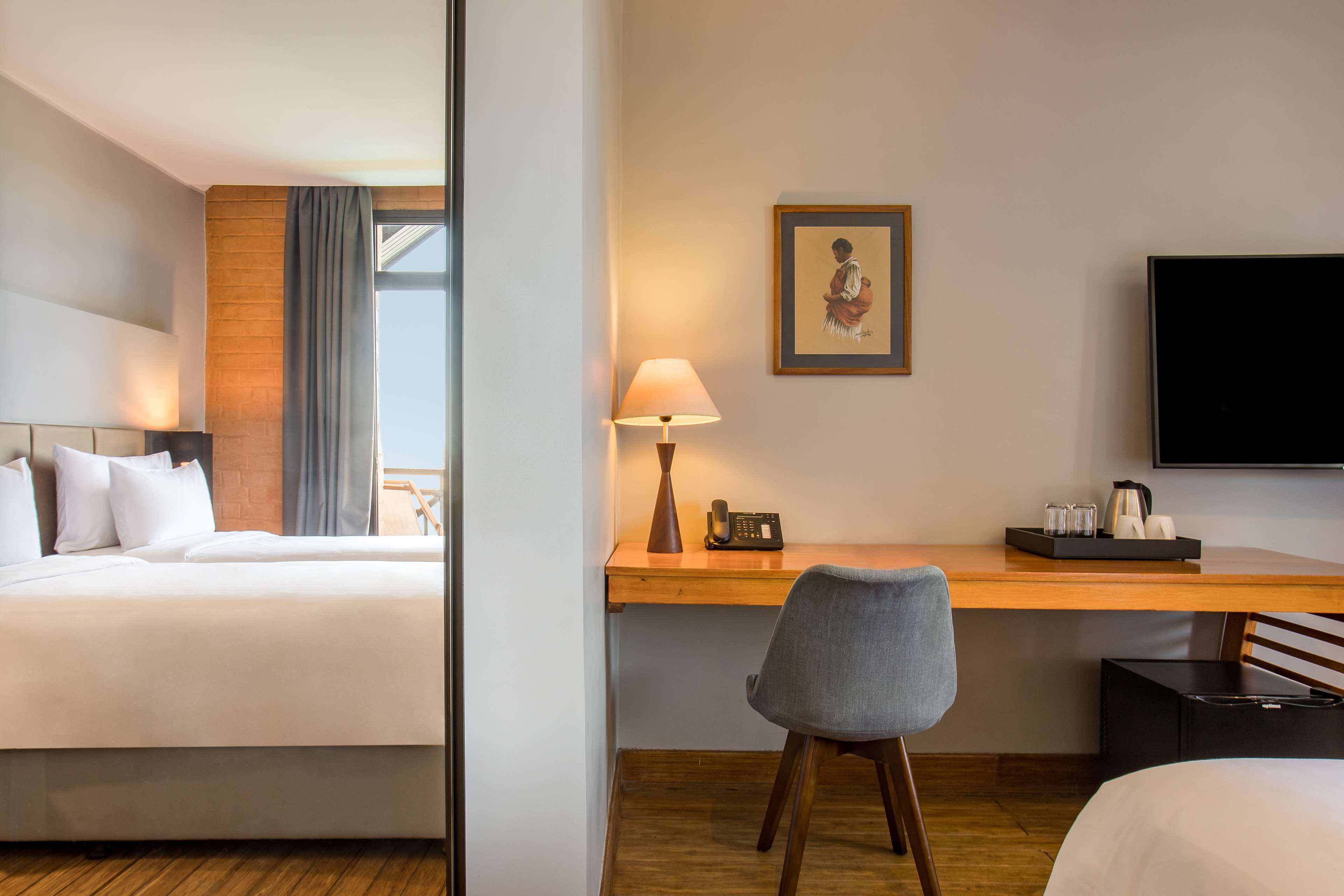 Quarto de hotel com uma cama, uma escrivaninha com luminária, uma cadeira, televisão e um quadro na parede.
