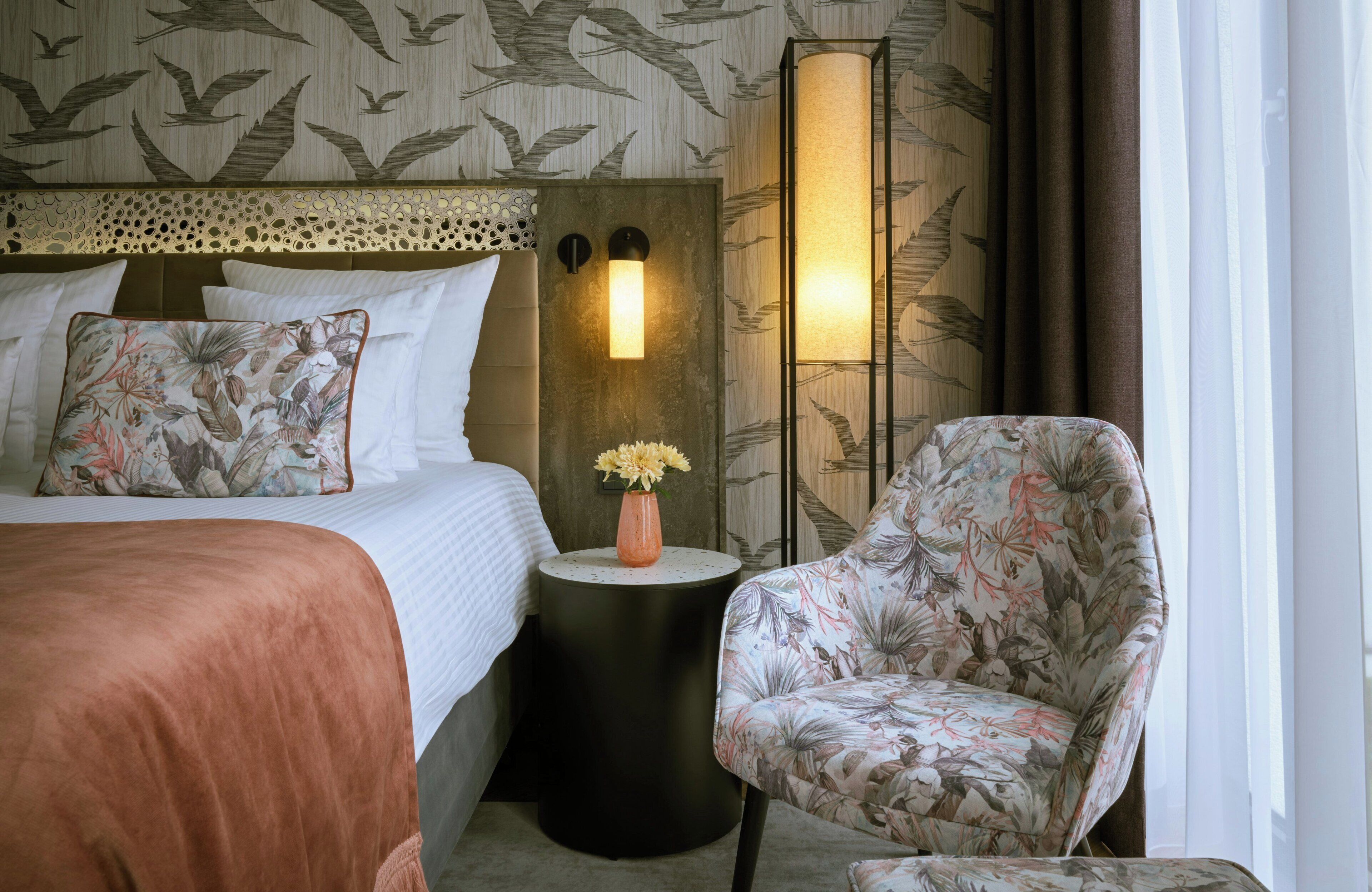 Quarto de hotel com cama, cadeira, mesa de cabeceira, luminária na parede e painel decorativo.