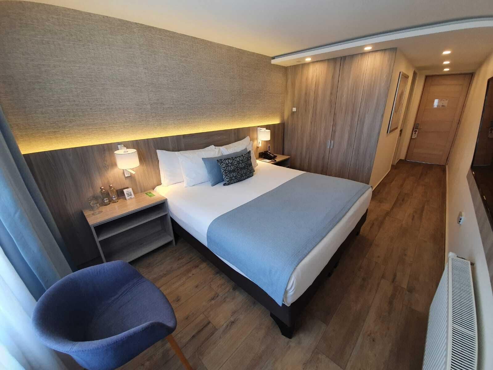 Quarto de hotel com cama grande, mesas de cabeceira, uma cadeira e armário embutido.
