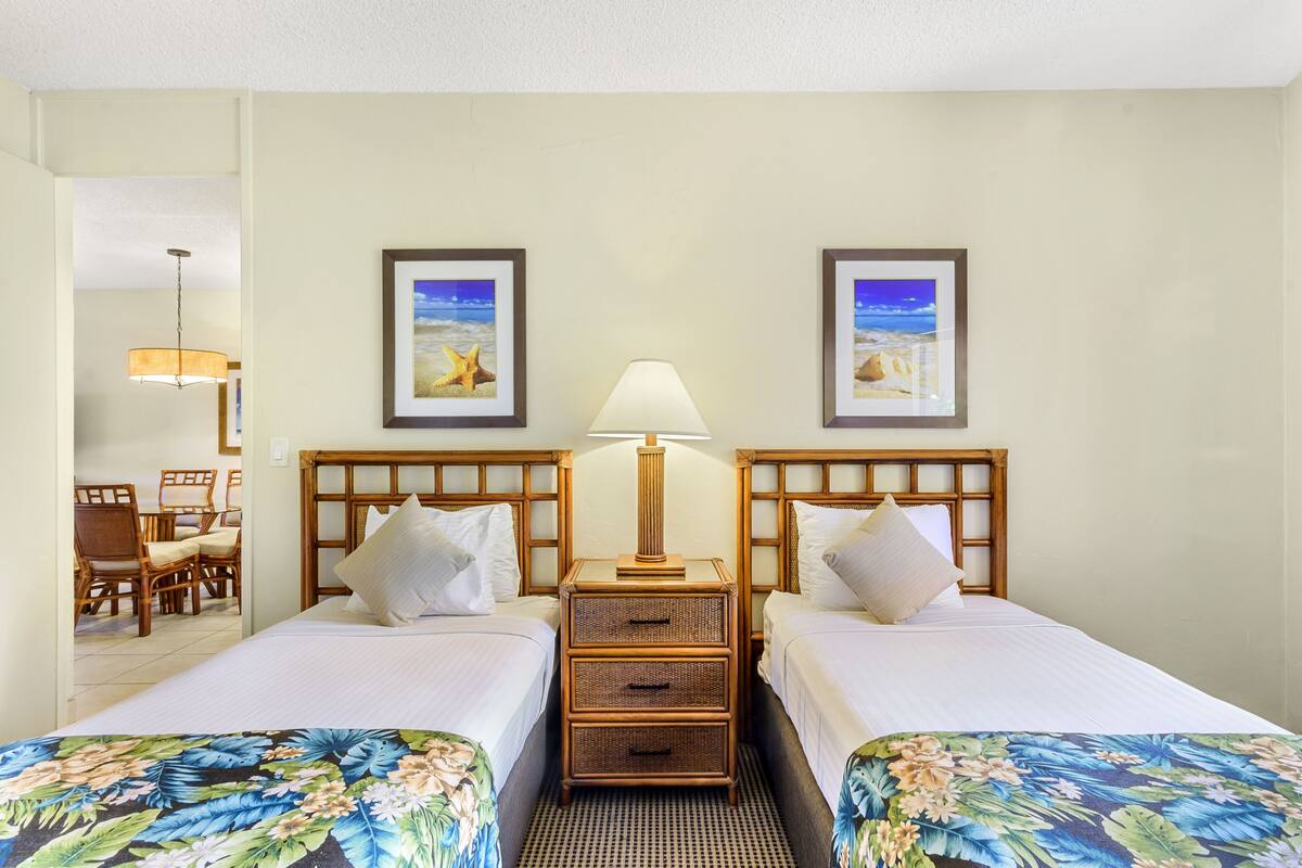 Quarto de hotel com duas camas, uma mesa de cabeceira com abajur, uma cadeira e quadros nas paredes.