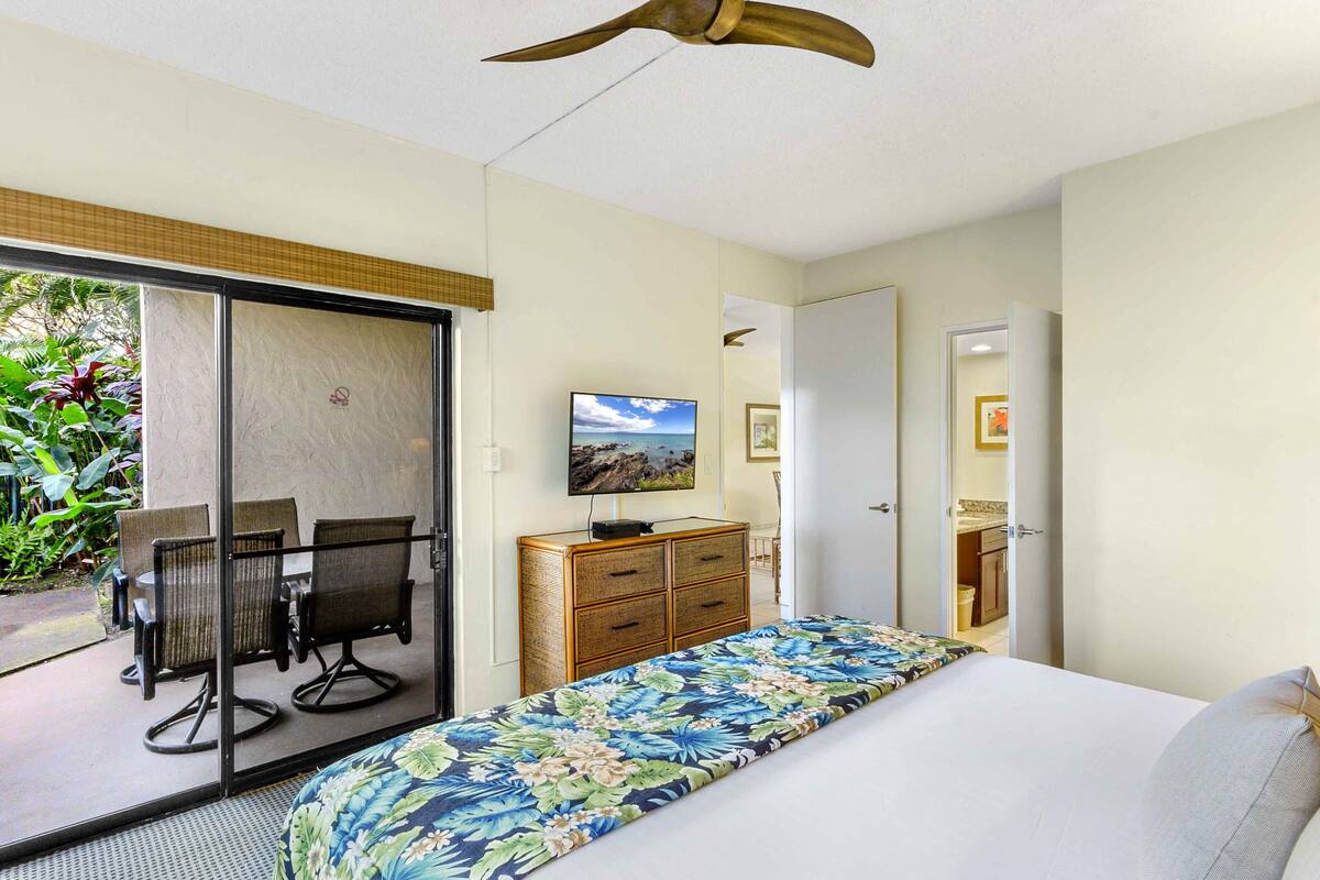 Quarto de hotel com uma cama, televisão, escrivaninha com cadeiras, cómoda de madeira e porta de vidro deslizante para área externa.