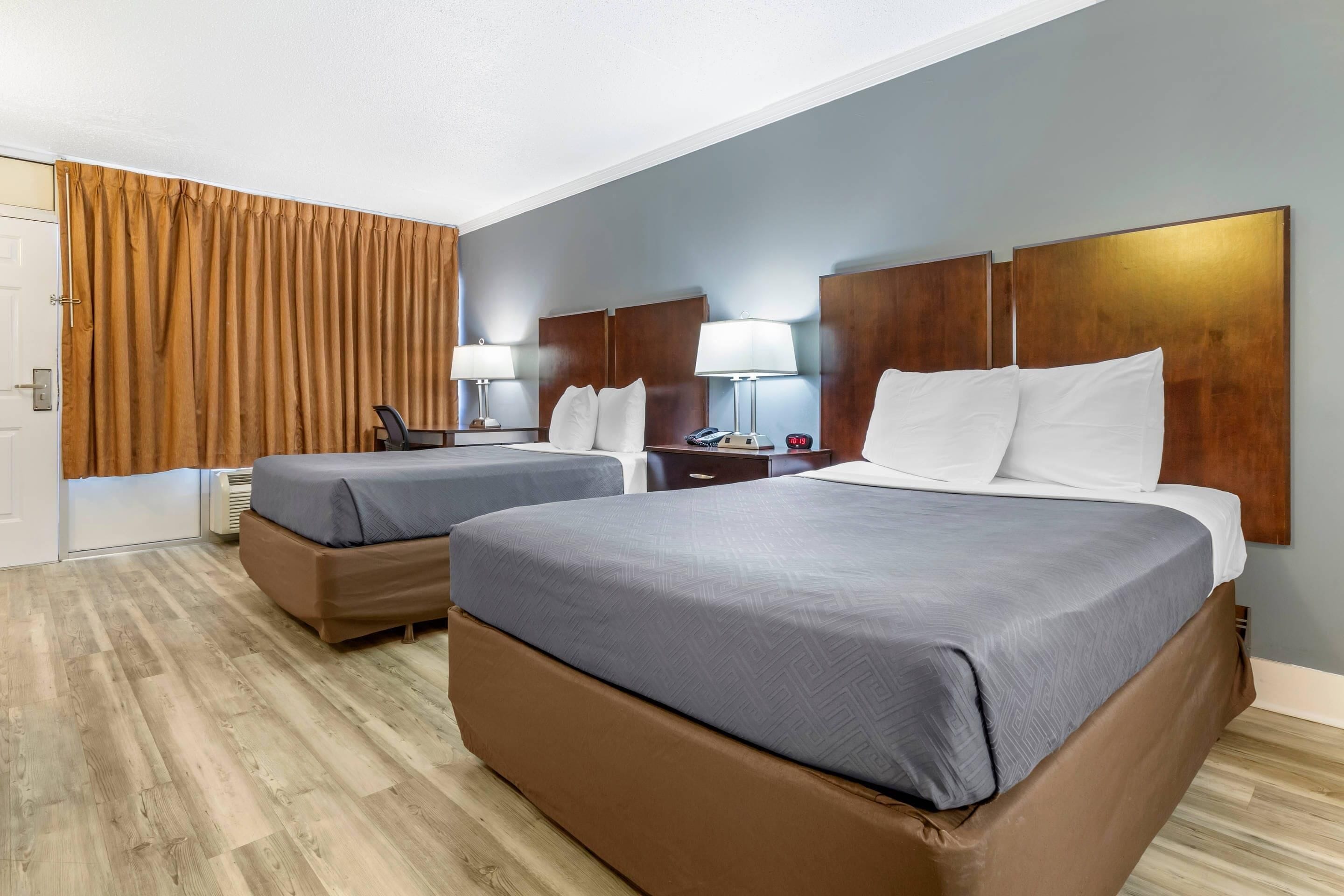 Quarto de hotel com duas camas, uma escrivaninha, luminárias e uma janela com cortinas.