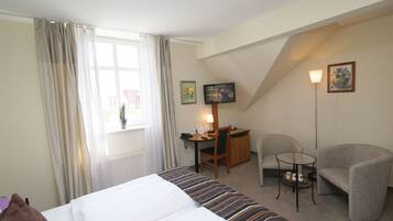 Quarto de hotel com cama, escrivaninha, duas cadeiras, mesa pequena e televisão.
