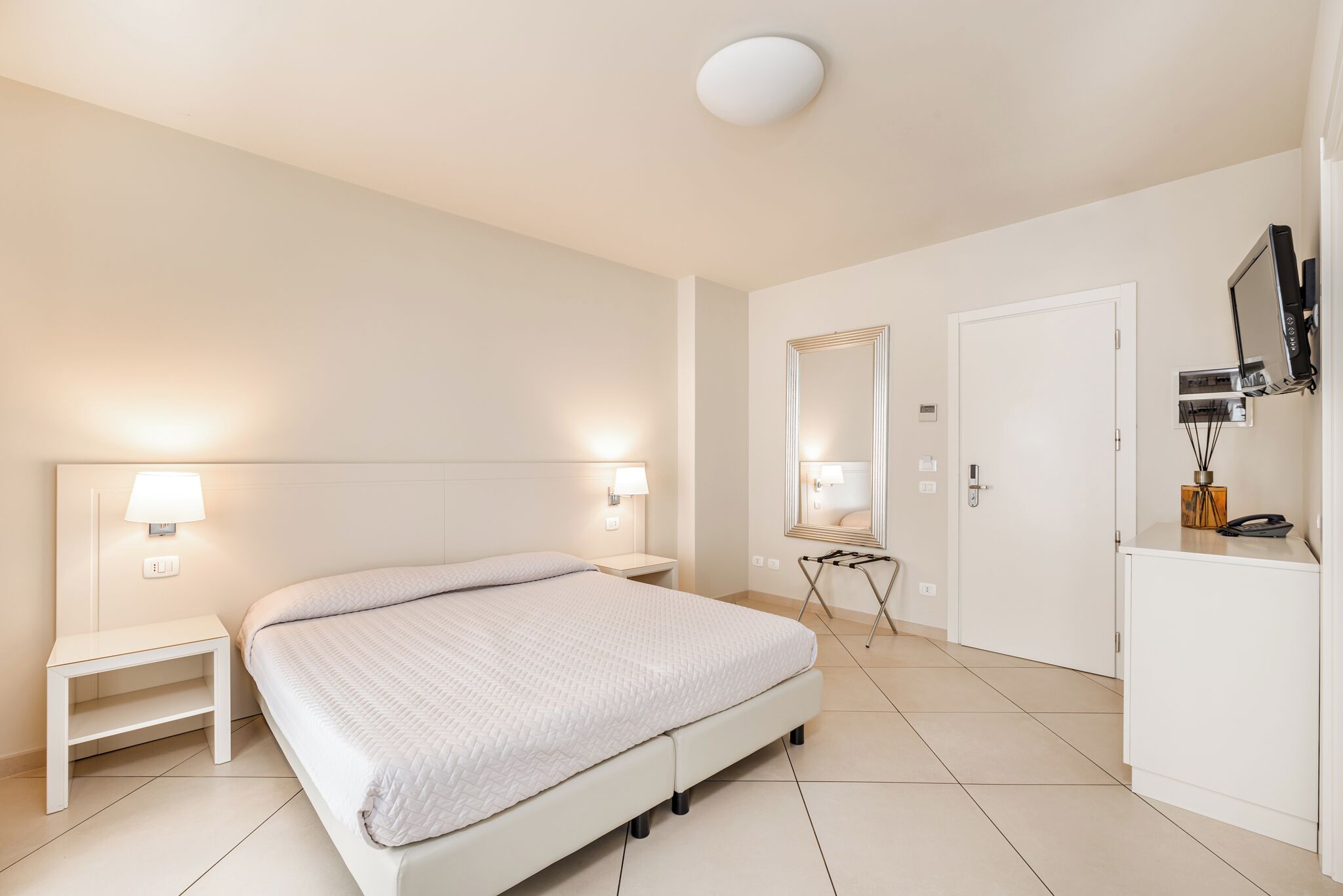 Quarto de hotel com uma cama, mesas de cabeceira, espelho, televisão e guarda-roupa.
