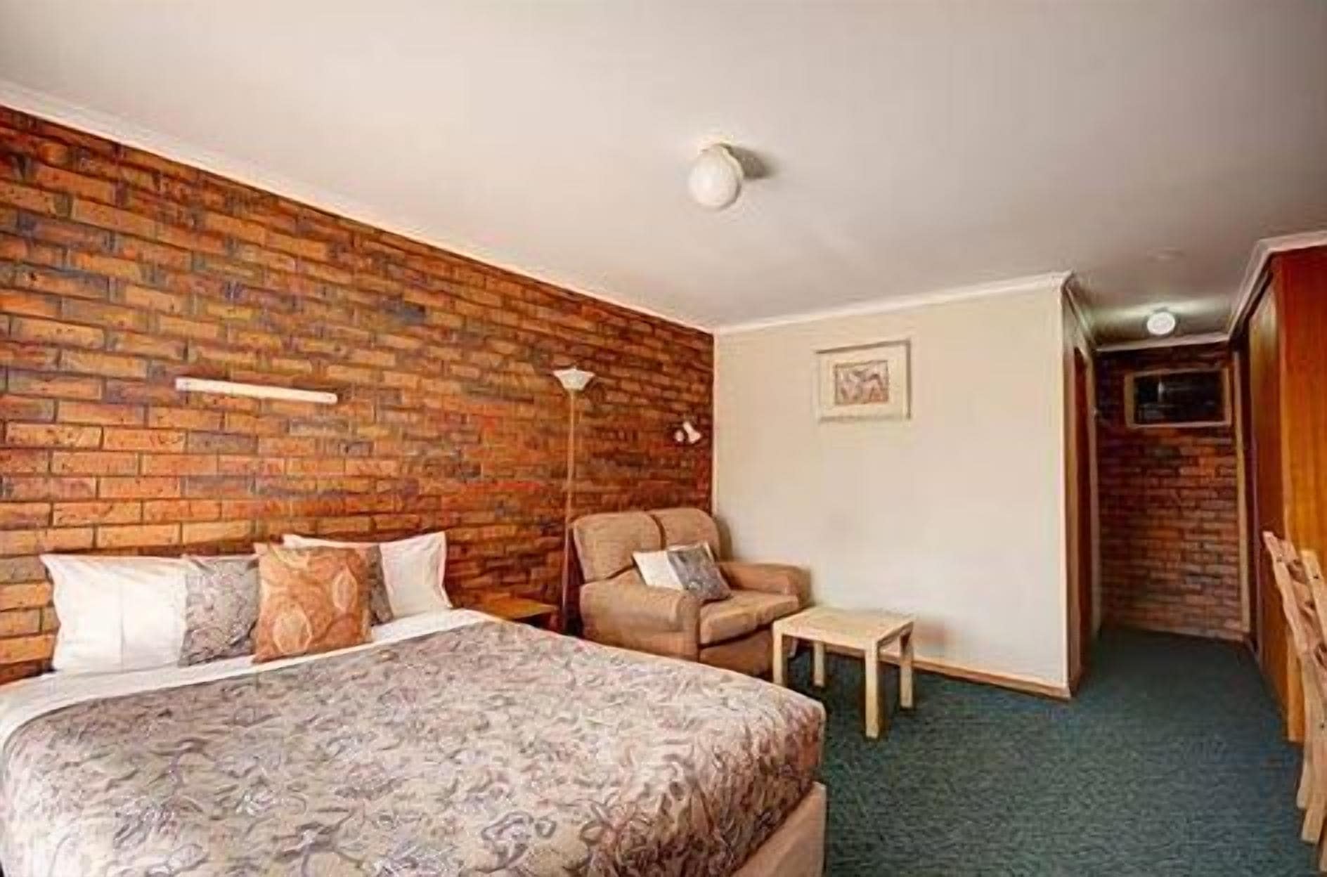 Quarto de hotel com parede de tijolos, uma cama, uma cadeira, uma pequena mesa e uma porta que dá para outro quarto.