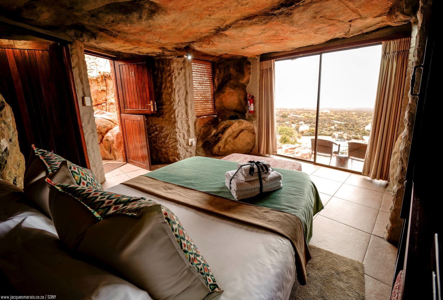 Quarto com cama, janela com vista, porta e parede de pedra.