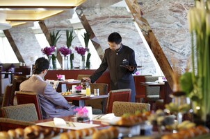 Interior de restaurante com parede de mármore, mesas, cadeiras e flores roxas em vasos.