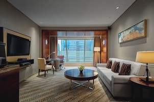 Quarto de hotel com televisão de tela plana, sofá, mesa de centro redonda e uma janela grande com vista para a cidade.
