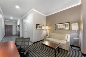 Quarto de hotel com sofá, mesa de centro, escrivaninha com cadeira, abajur e quadros nas paredes.