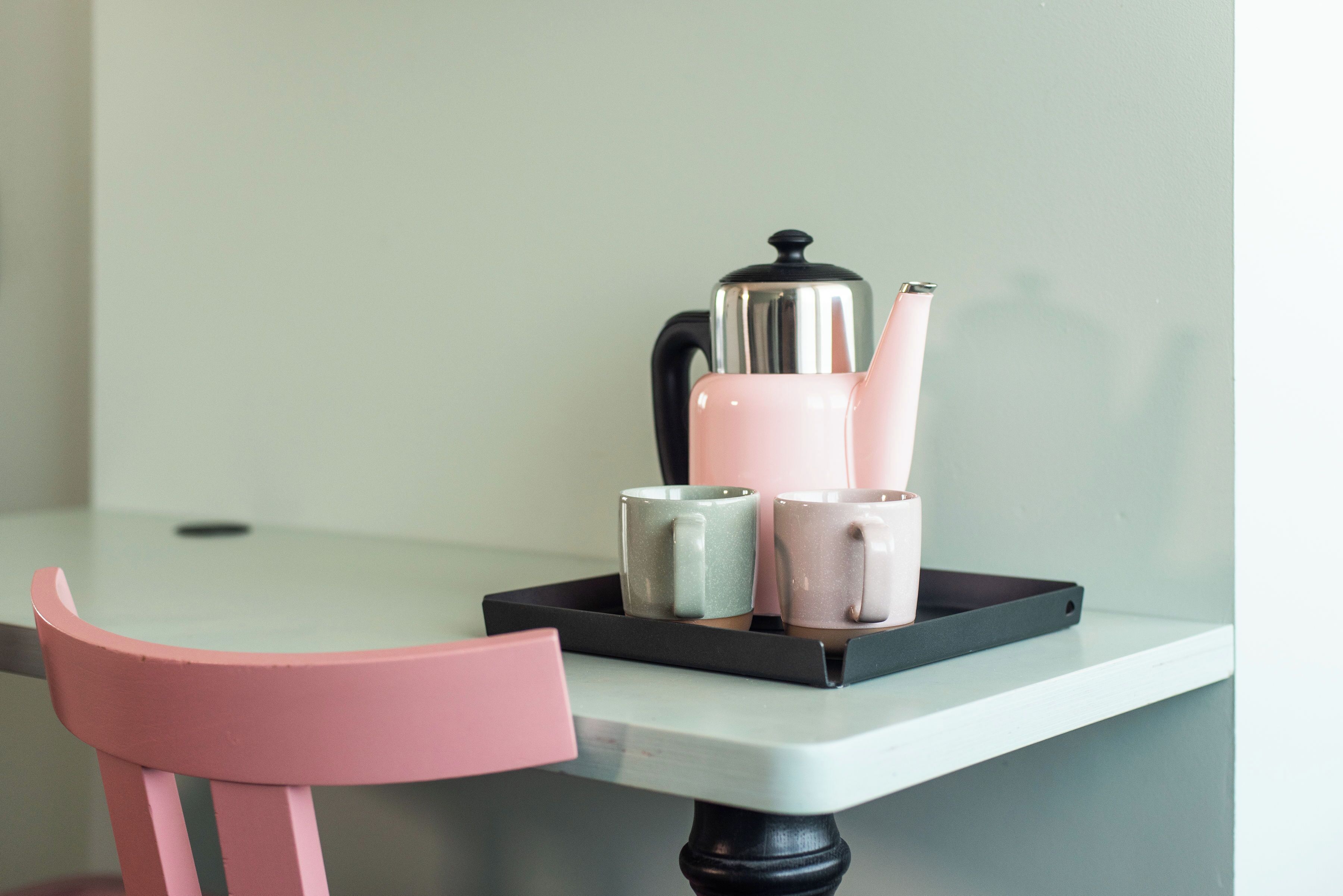 Uma mesa com uma bandeja que contém um bule rosa, duas chávenas e um pequeno recipiente.