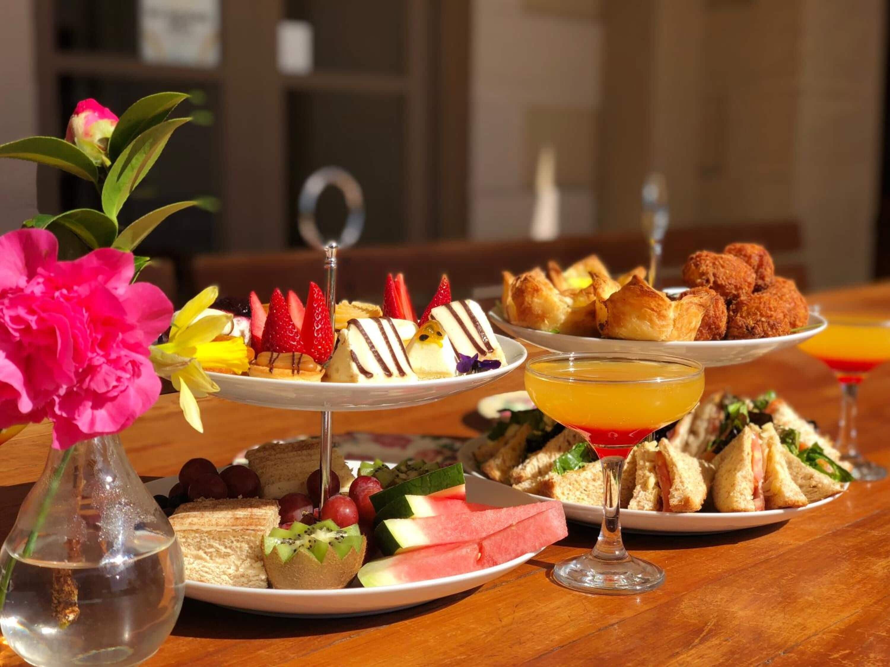 Uma mesa com diversos alimentos, incluindo sanduíches, doces e frutas, além de um vaso de flores e um copo de suco de laranja.