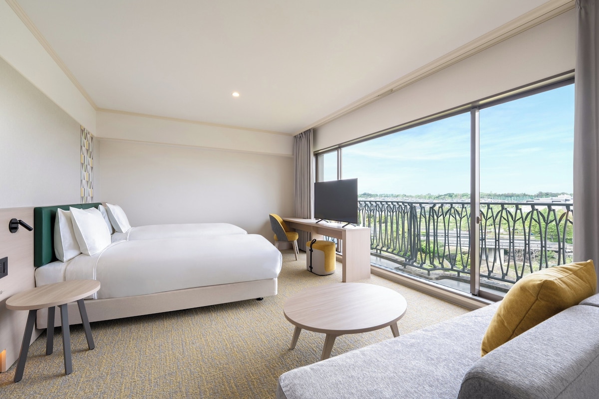 Quarto de hotel com uma cama, uma escrivaninha com televisão, um sofá e uma janela grande com vista.