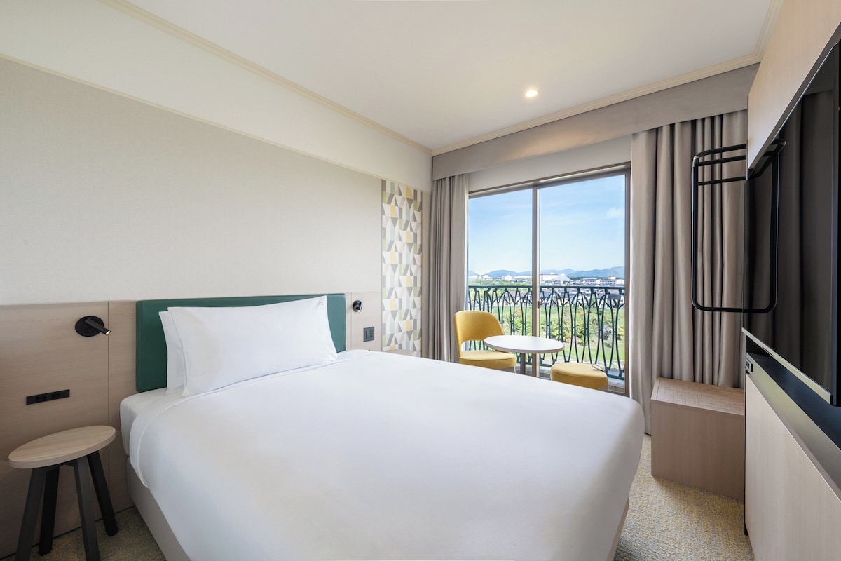 Quarto de hotel com uma cama grande, uma mesa redonda pequena, uma cadeira, televisão e varanda com vista.